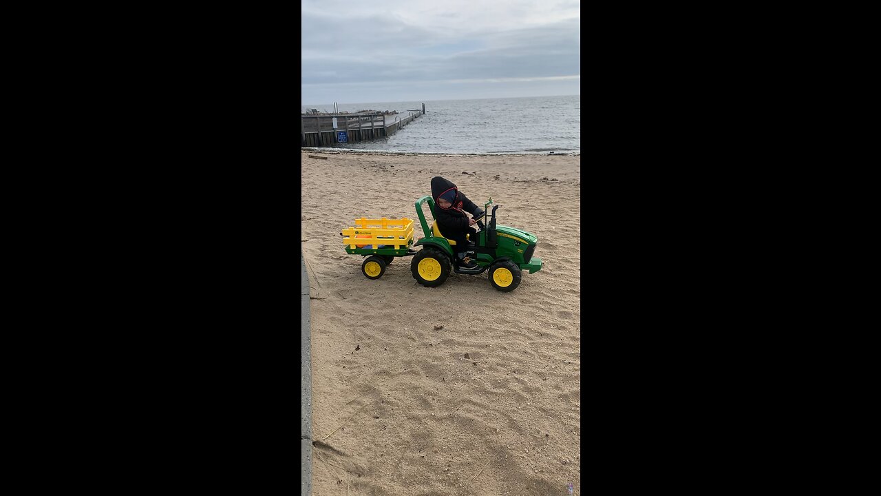 2 year old driving John Deere tractor across beach on sand to the park ...